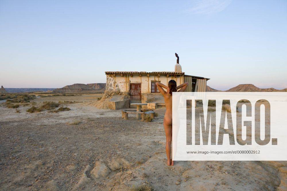 Nude young girl standing in desert in front of a barrack